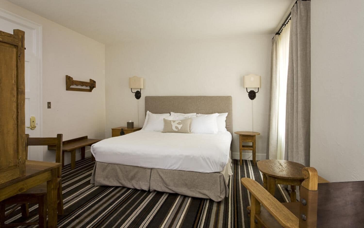 Quarto de hotel com uma cama grande, duas mesas de cabeceira, um banco de madeira, um tapete listrado e vista para o exterior.