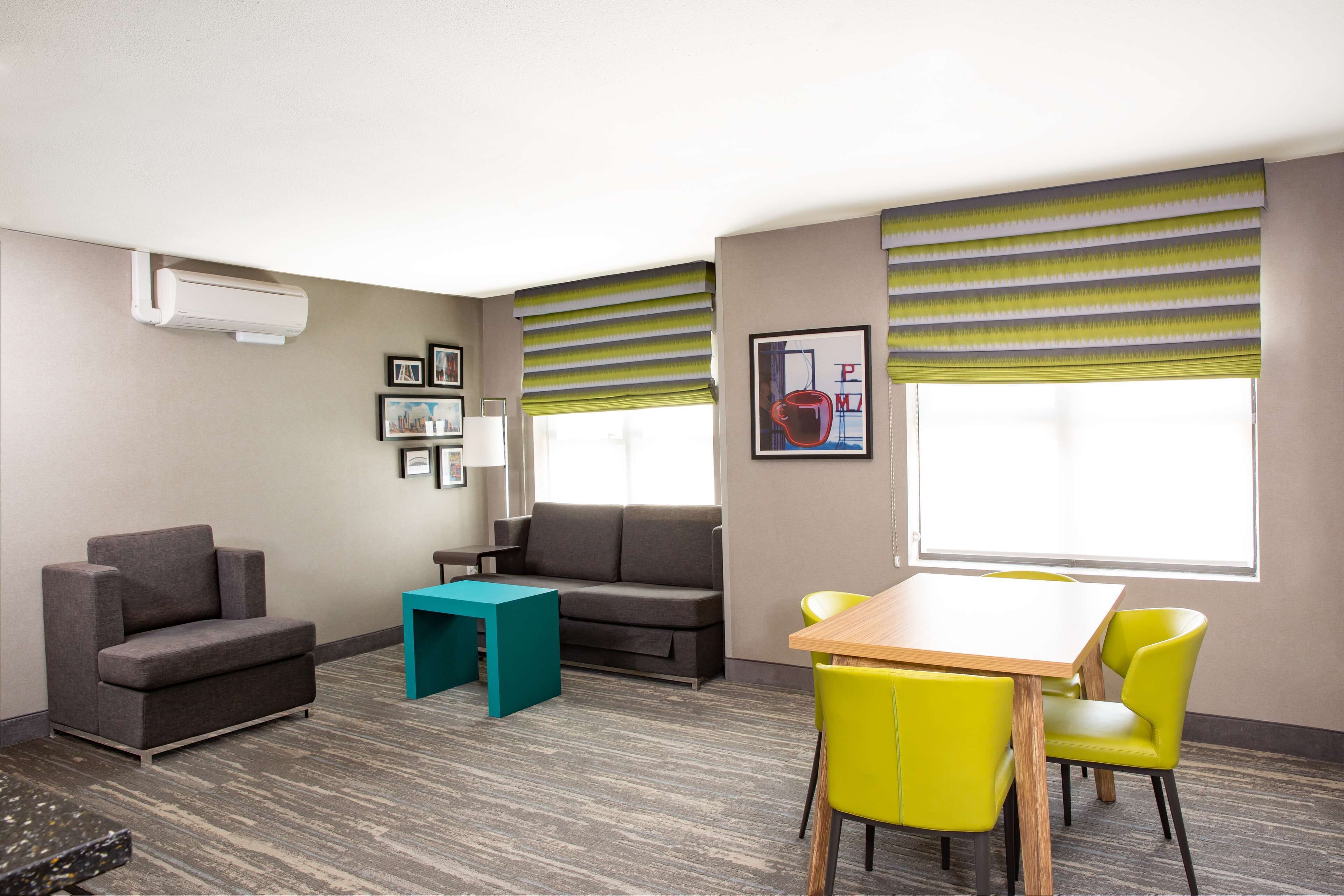 Quarto de hotel moderno com sofá, uma mesa pequena, uma mesa de jantar com cadeiras e janelas amplas com persianas listradas.