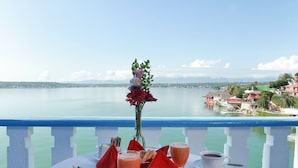 Mesa posta com itens de café da manhã, incluindo panquecas, frutas e café, com vista para um corpo d'água e edifícios ao fundo.