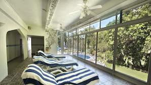 Quarto iluminado pelo sol, com ventiladores de teto, janelas amplas e toalhas listradas em espreguiçadeiras.