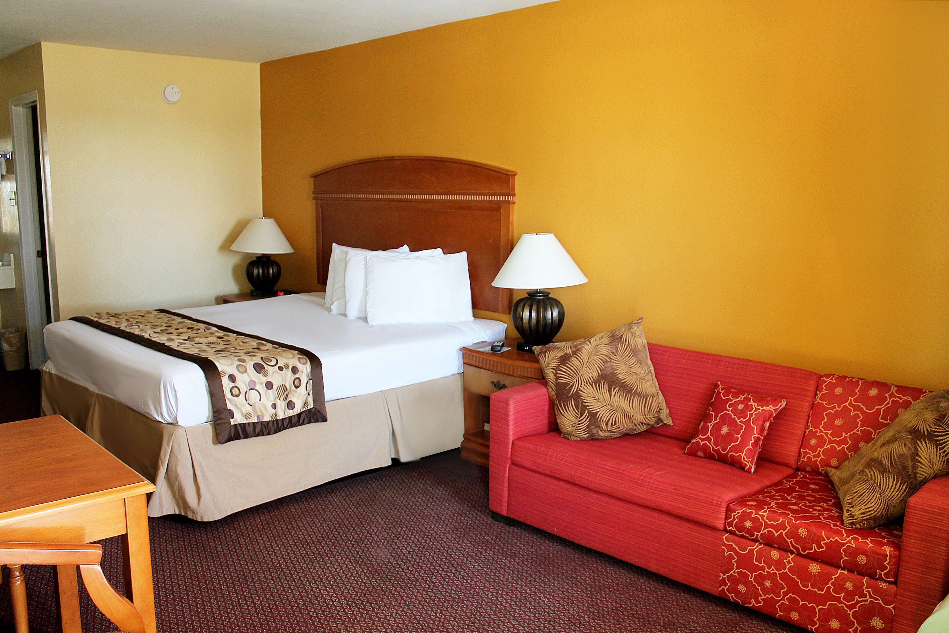 Quarto de hotel com uma cama, um sofá vermelho, duas mesas de cabeceira com luminárias e uma mesa de cabeceira de madeira.