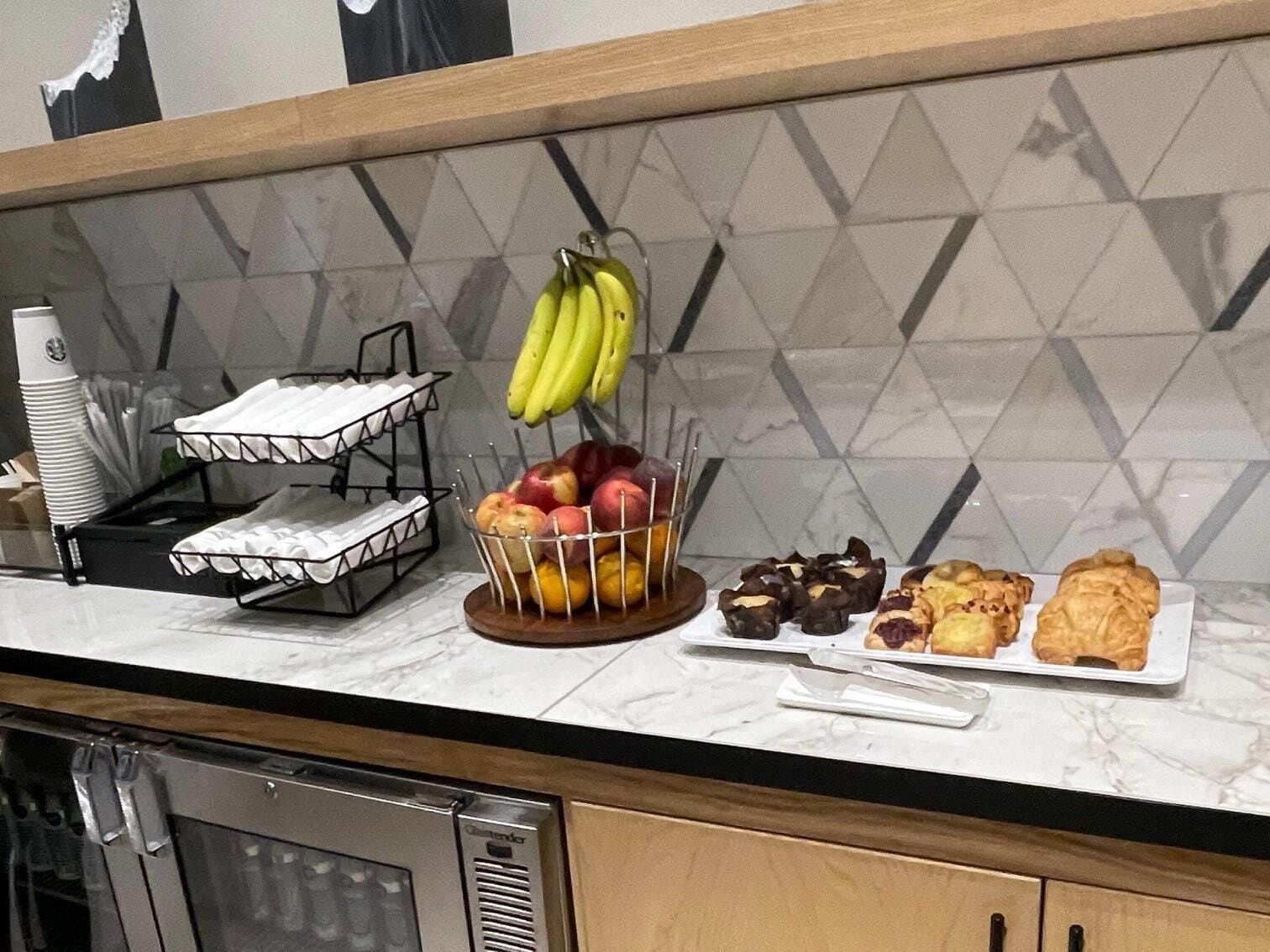 Uma bancada de cozinha com uma cesta de frutas, um suporte para bananas, uma bandeja de ovos e um prato de doces.