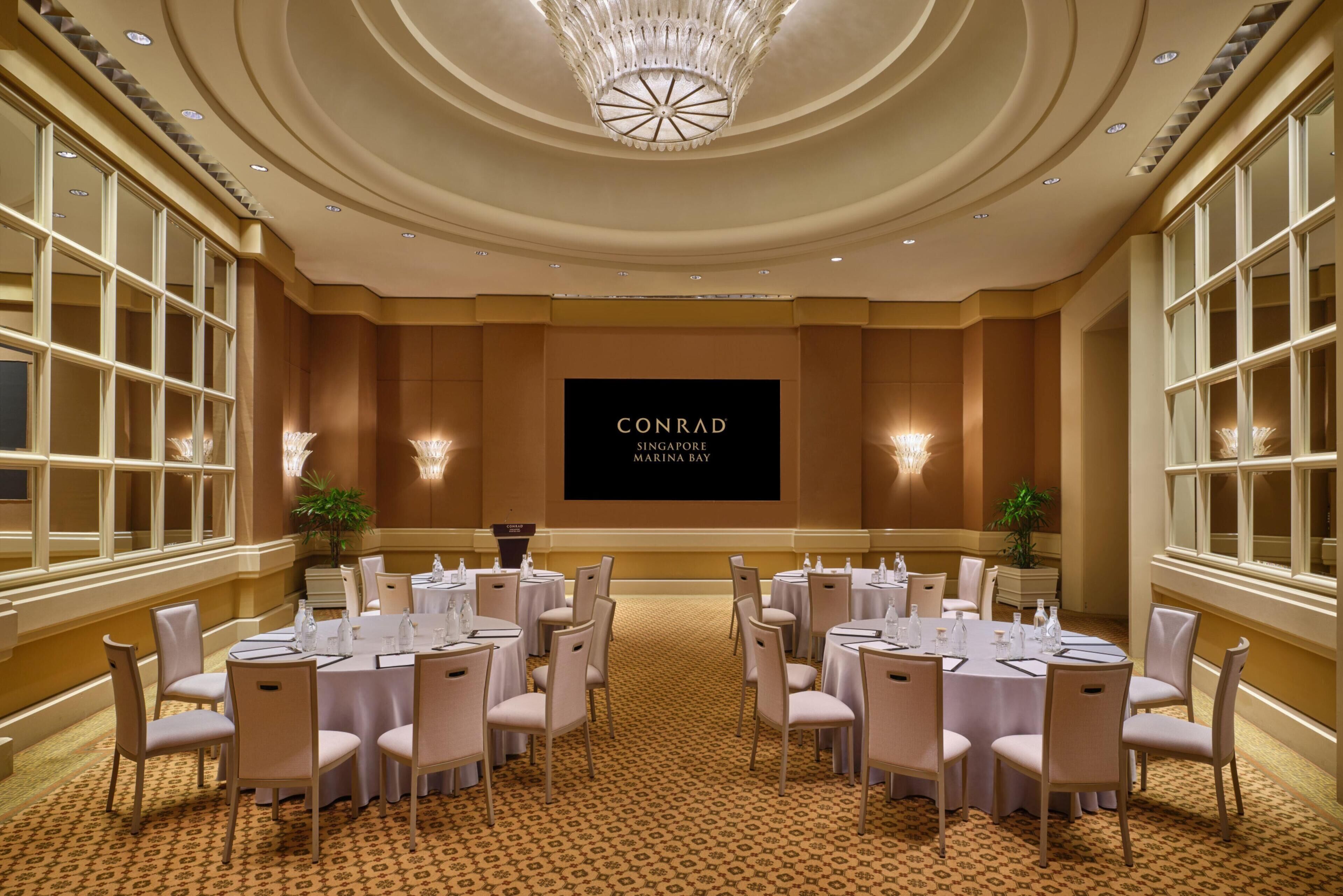Uma sala de conferências com mesas redondas, cadeiras, um candelabro e uma grande tela exibindo 'CONRAD SINGAPORE MARINA BAY'.