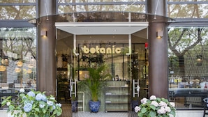 Entrada de um edifício moderno com fachada de vidro, uma placa com a palavra "botanic" e plantas em vasos na calçada.