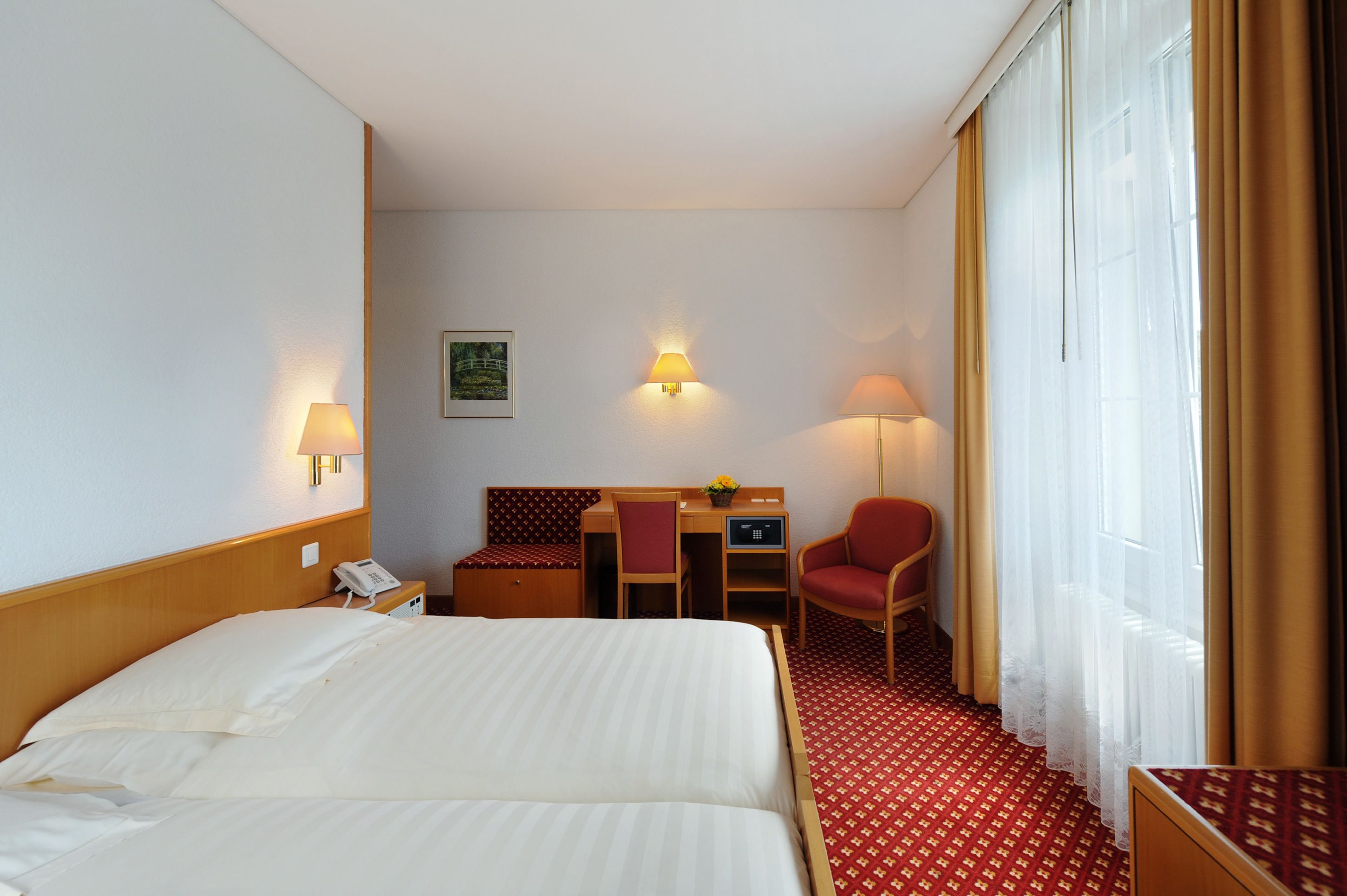 Quarto de hotel com cama, escrivaninha, cadeira e duas luminárias fixadas na parede.