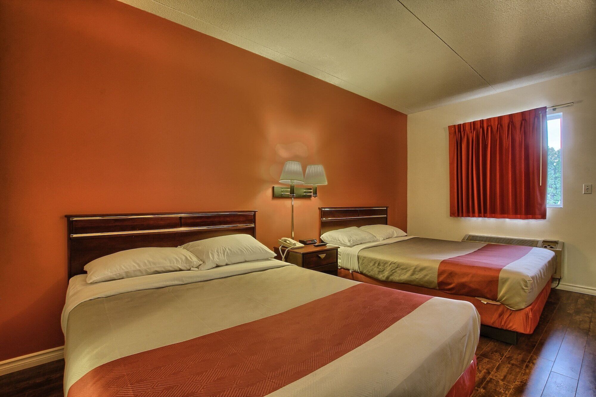 Quarto de hotel com duas camas, uma mesa de cabeceira com abajur e uma janela com cortinas vermelhas.