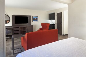 Quarto de hotel com cama, escrivaninha com TV de tela plana, poltrona vermelha e mesa pequena.