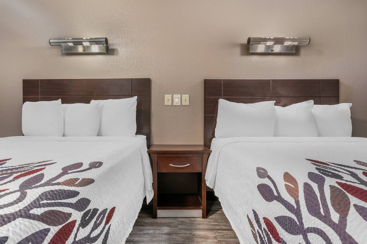 Quarto de hotel com duas camas, uma mesa de cabeceira de madeira e arandelas na parede.