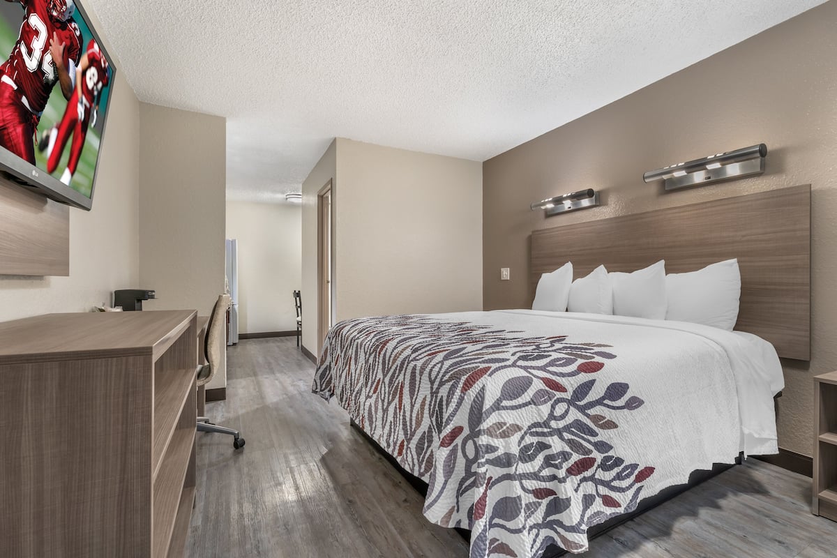 Quarto de hotel com cama, escrivaninha, televisão e cabeceira iluminada.