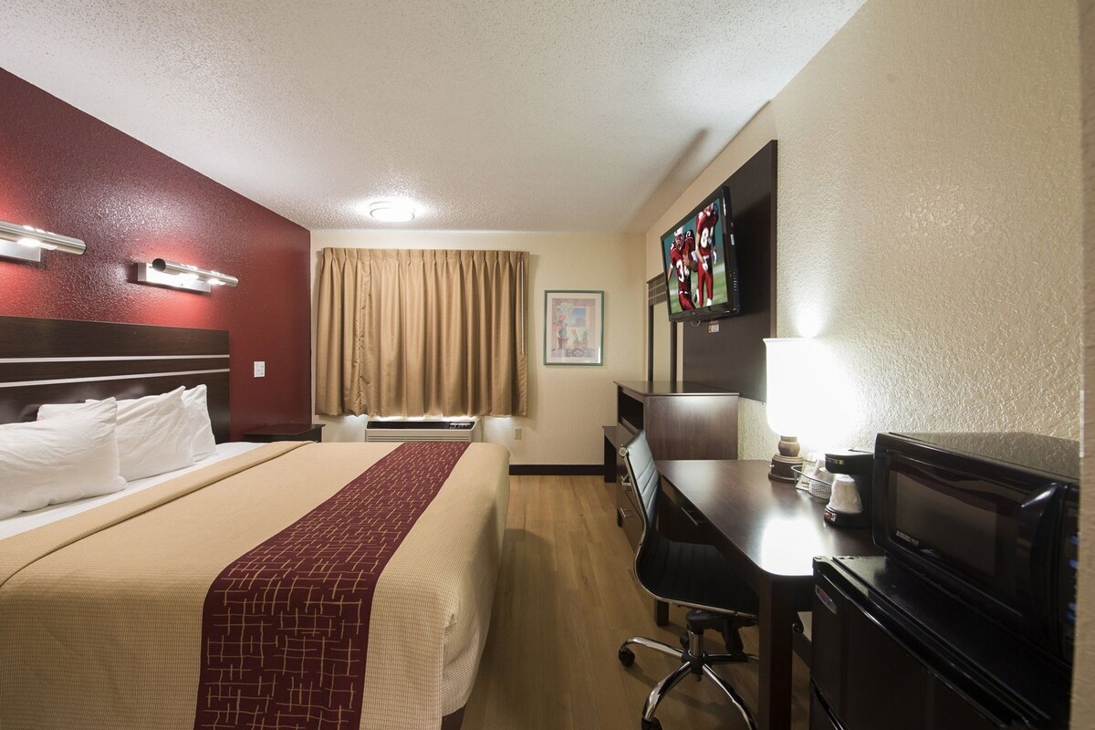 Quarto de hotel com cama, escrivaninha, cadeira, televisão e janela com cortinas.
