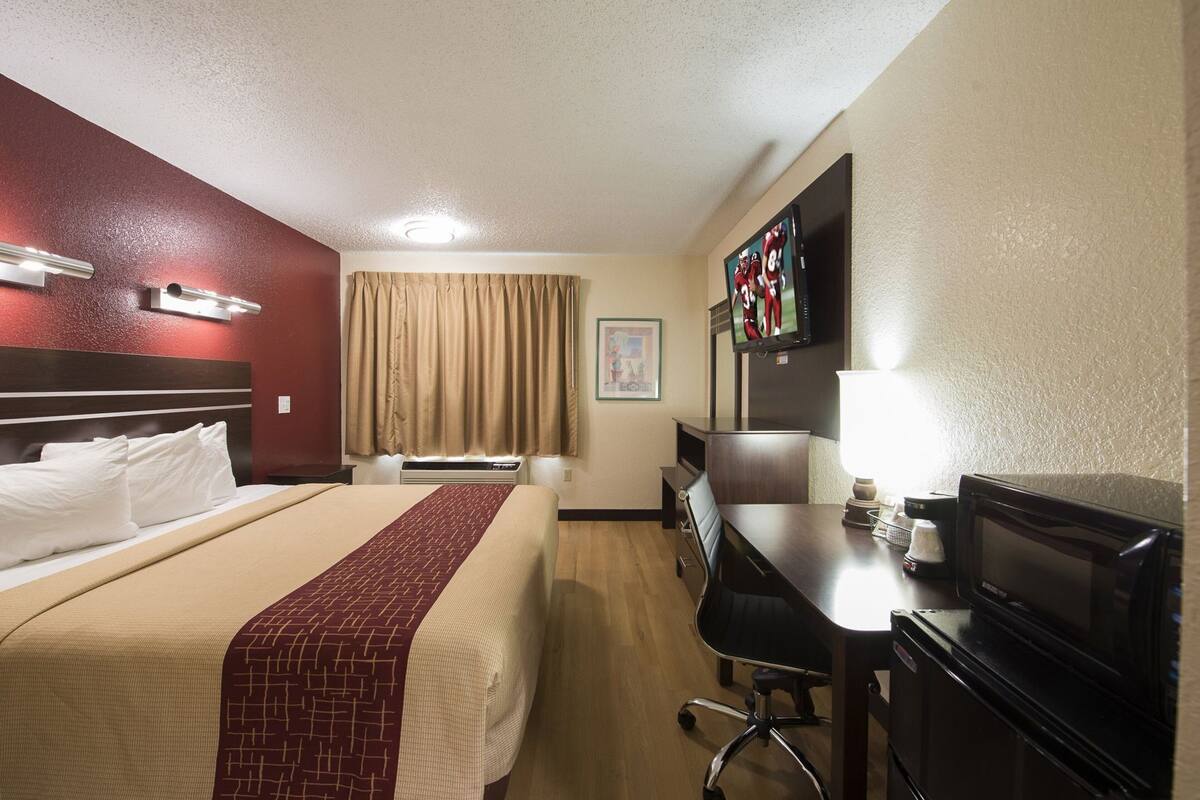 Quarto de hotel com cama, escrivaninha, cadeira, televisão e janela com cortinas.