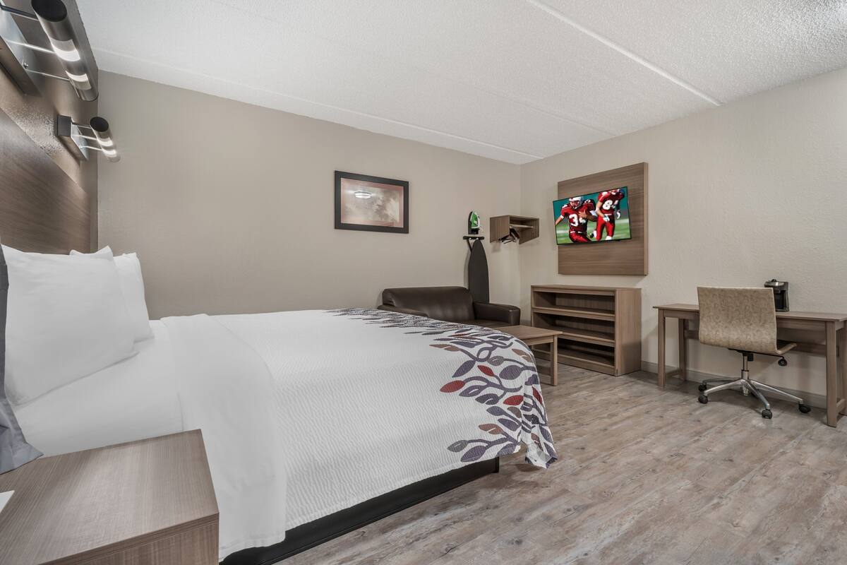 Quarto de hotel com cama, escrivaninha, cadeira, televisão e um quadro na parede.