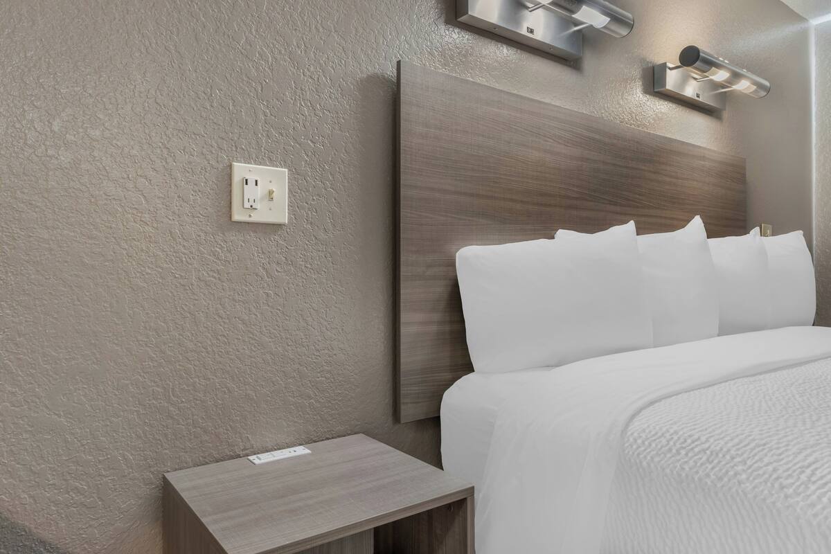 Quarto de hotel com parede texturizada, cabeceira de madeira, mesa de cabeceira e luminária fixa na parede.