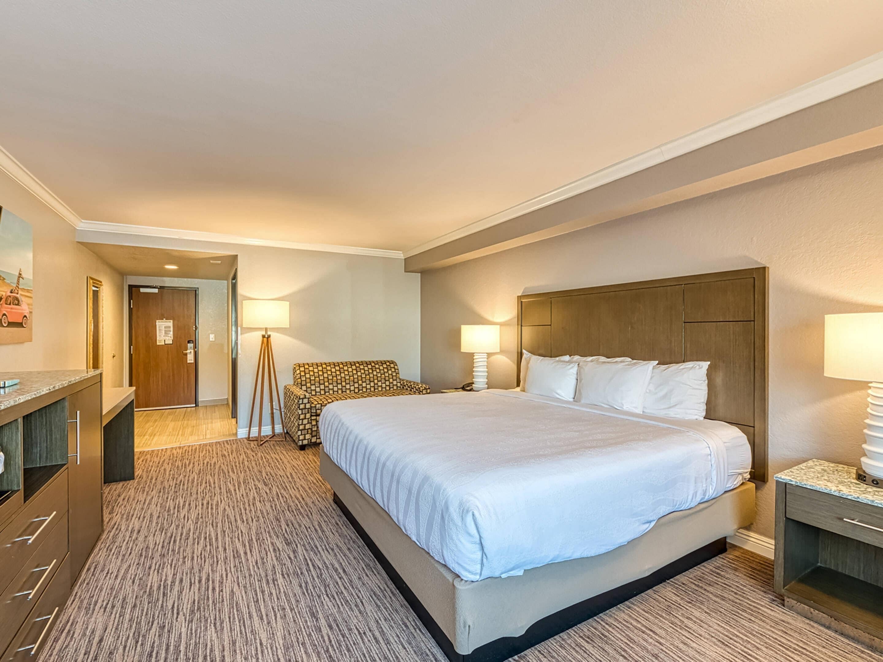 Quarto de hotel com uma cama grande, uma escrivaninha, uma cadeira e uma mesa de cabeceira com luminárias.