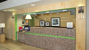 Balcão de recepção de hotel com bancada de granito, quadros decorativos, placa de boas-vindas e uma luminária.