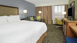 Quarto de hotel com uma cama grande, uma escrivaninha com televisão, uma cadeira e um abajur.