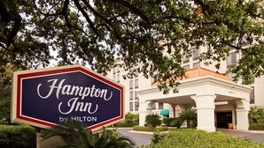 Placa de um Hampton Inn by Hilton em frente a um edifício com fachada branca e telhado vermelho.