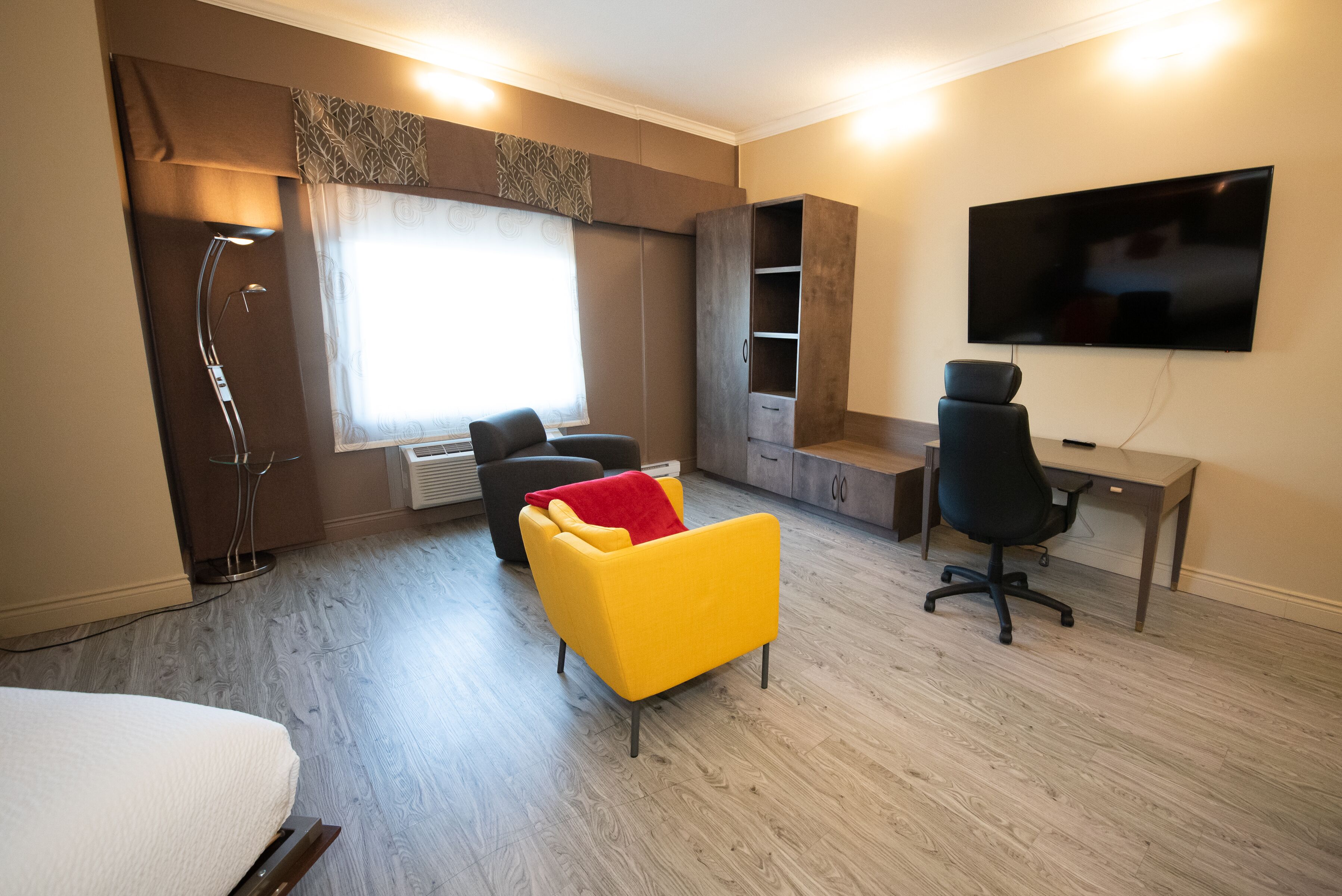 Quarto moderno com poltrona amarela, cadeira de escritório preta, televisão de tela plana e uma janela grande com cortinas.