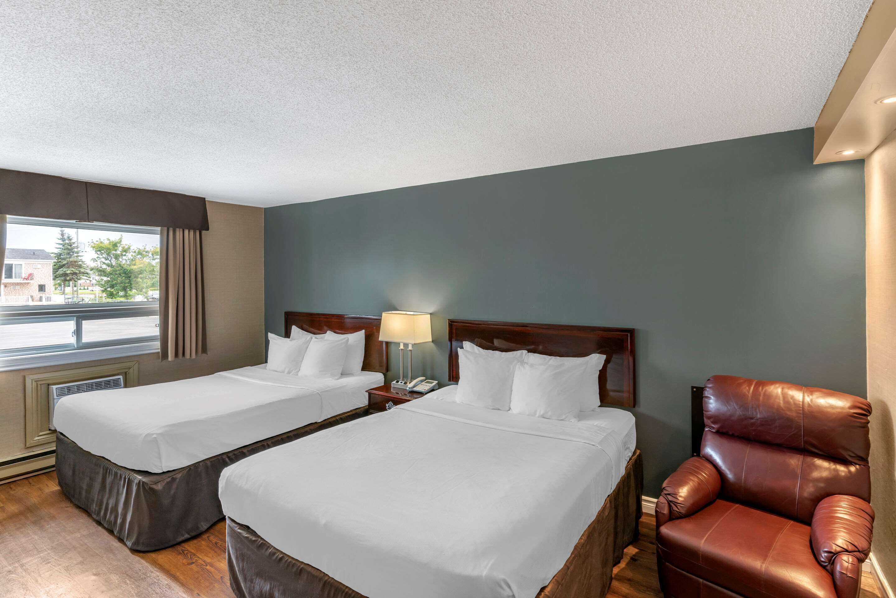 Quarto de hotel com duas camas, uma poltrona de couro, mesa de cabeceira com abajur e uma janela com cortinas.