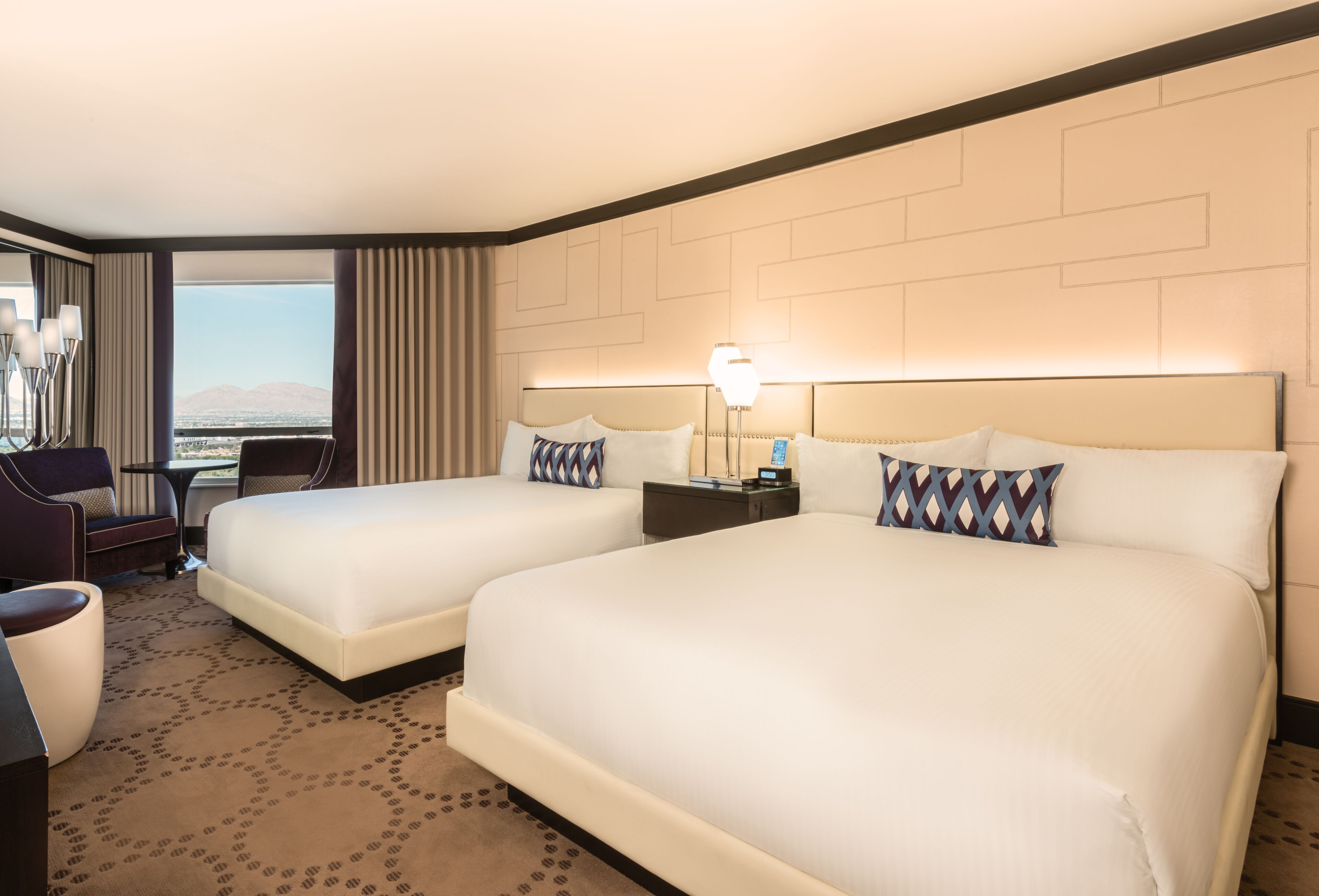 Quarto de hotel com duas camas, uma escrivaninha, uma cadeira e uma janela ampla com vista.