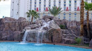 Piscina com cascata, cercada por grandes pedras e palmeiras, com um prédio de hotel ao fundo.