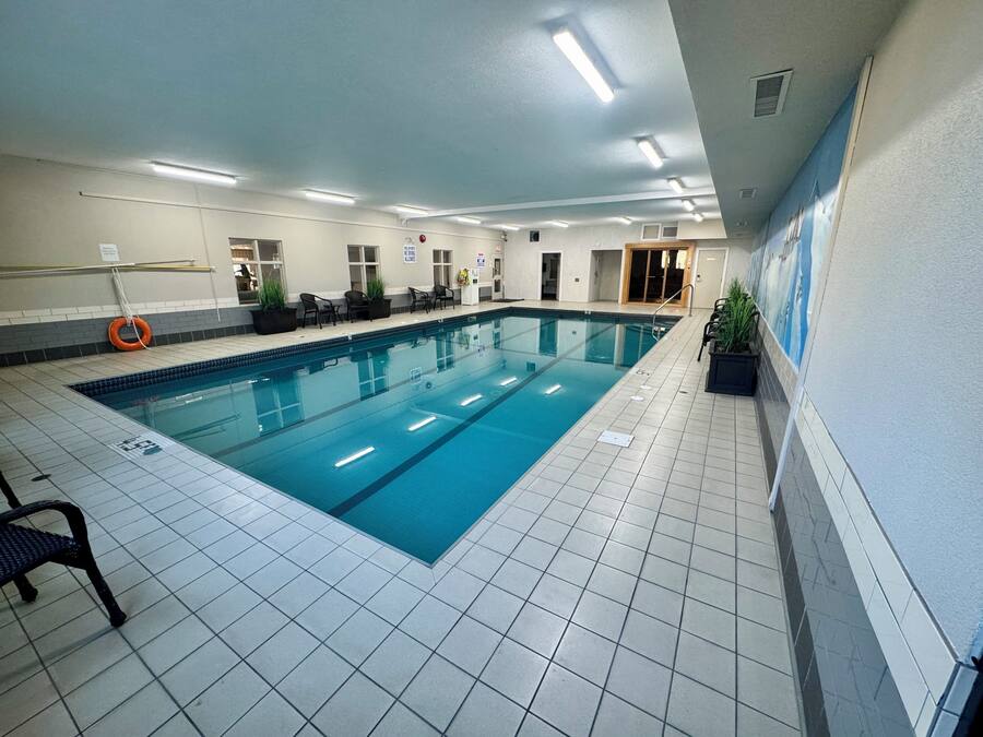 Piscina interior com chão ladrilhado, um salva-vidas e cadeiras na lateral.