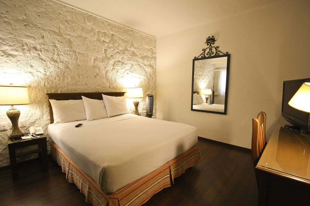 Quarto com parede de pedra, cama com roupa de cama branca, duas luminárias de cabeceira, um espelho e uma mesa de cabeceira de madeira.