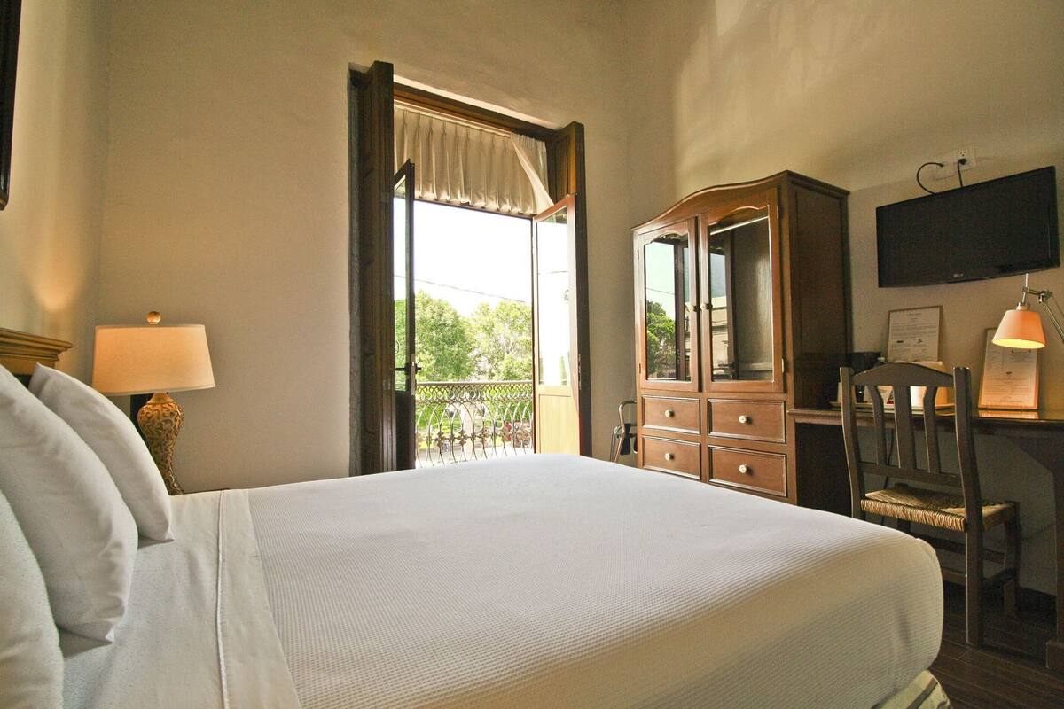 Quarto de hotel com cama, uma escrivaninha com cadeira, televisão e janela com vista para vegetação.