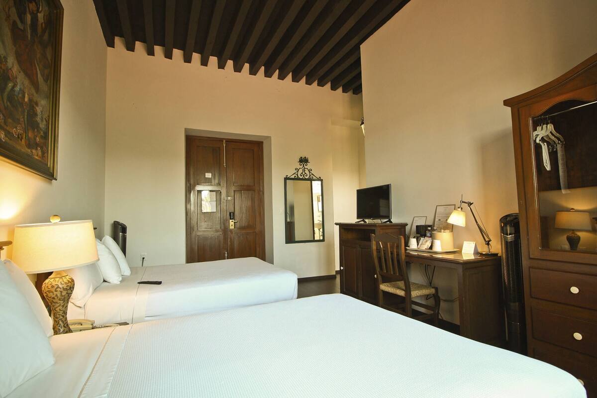 Quarto de hotel com duas camas, uma escrivaninha com computador, uma cadeira, penteadeira, espelho e um quadro na parede.