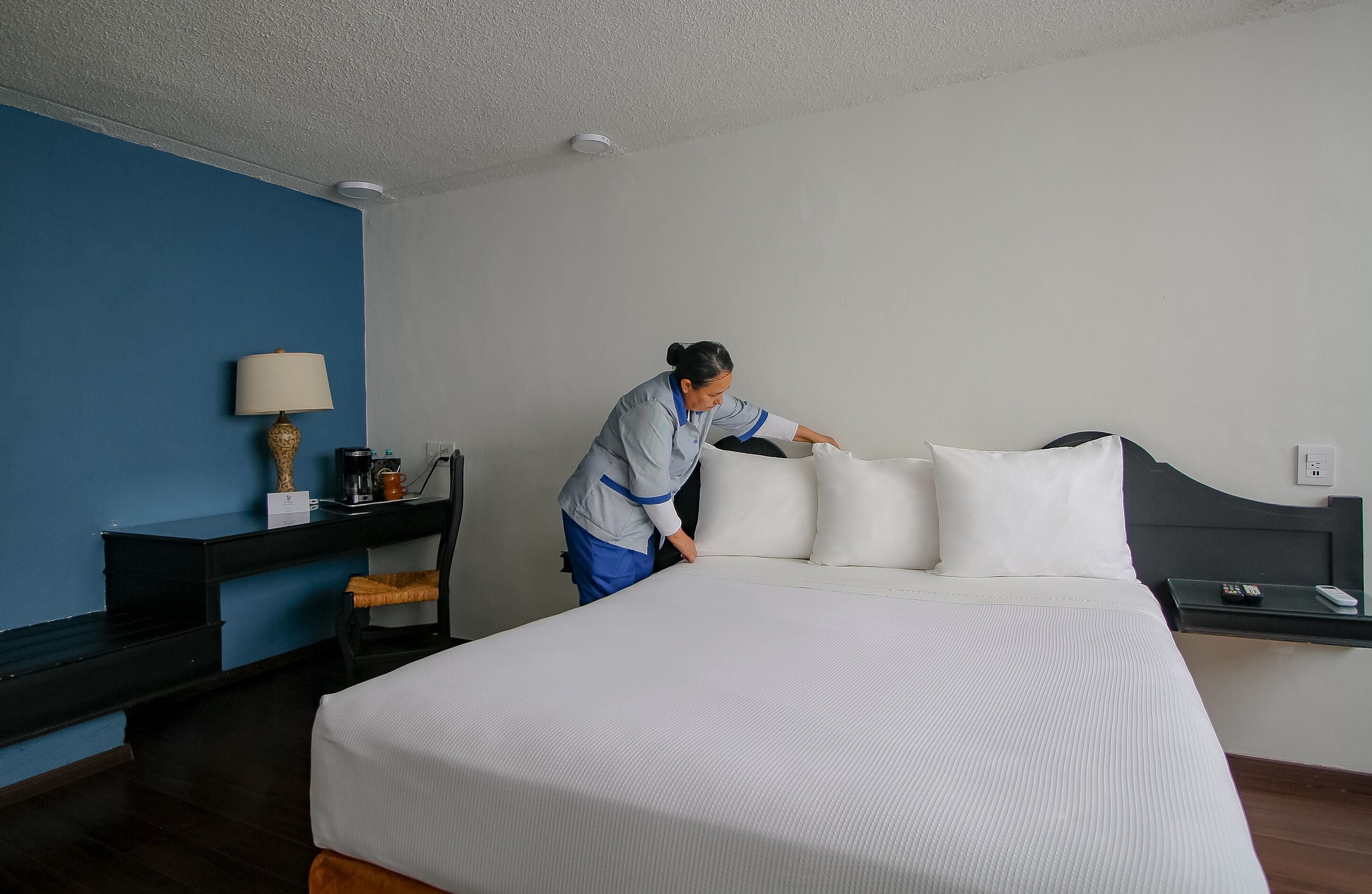 Quarto de hotel com uma cama, mesa de cabeceira, luminária, cafeteira e parede azul.