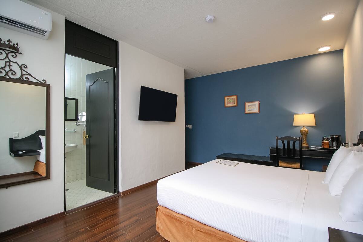 Quarto de hotel com uma cama, uma escrivaninha com abajur, uma televisão e um banheiro visível por uma porta aberta.