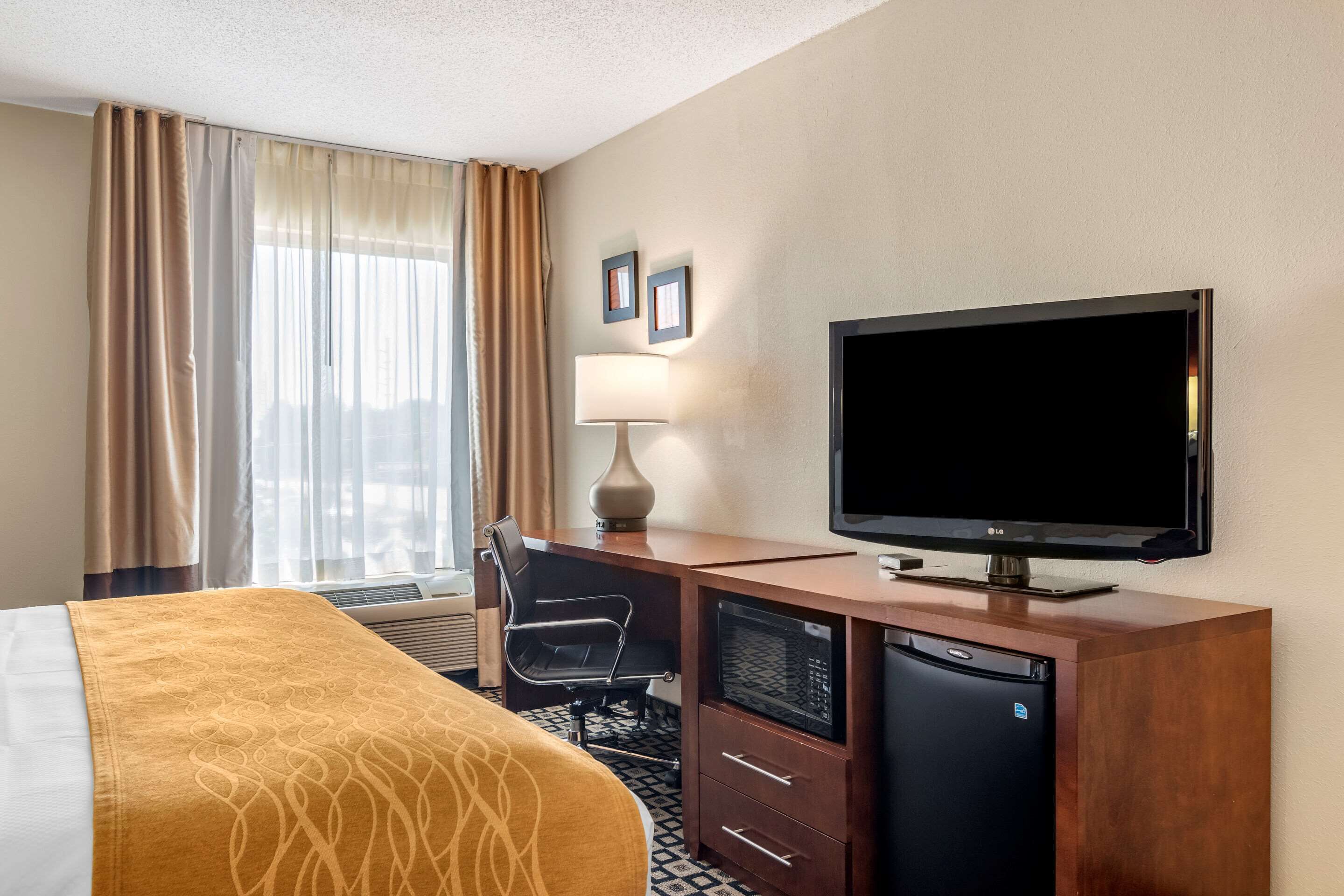Quarto de hotel com cama, escrivaninha, televisão, frigorífico e cadeira.