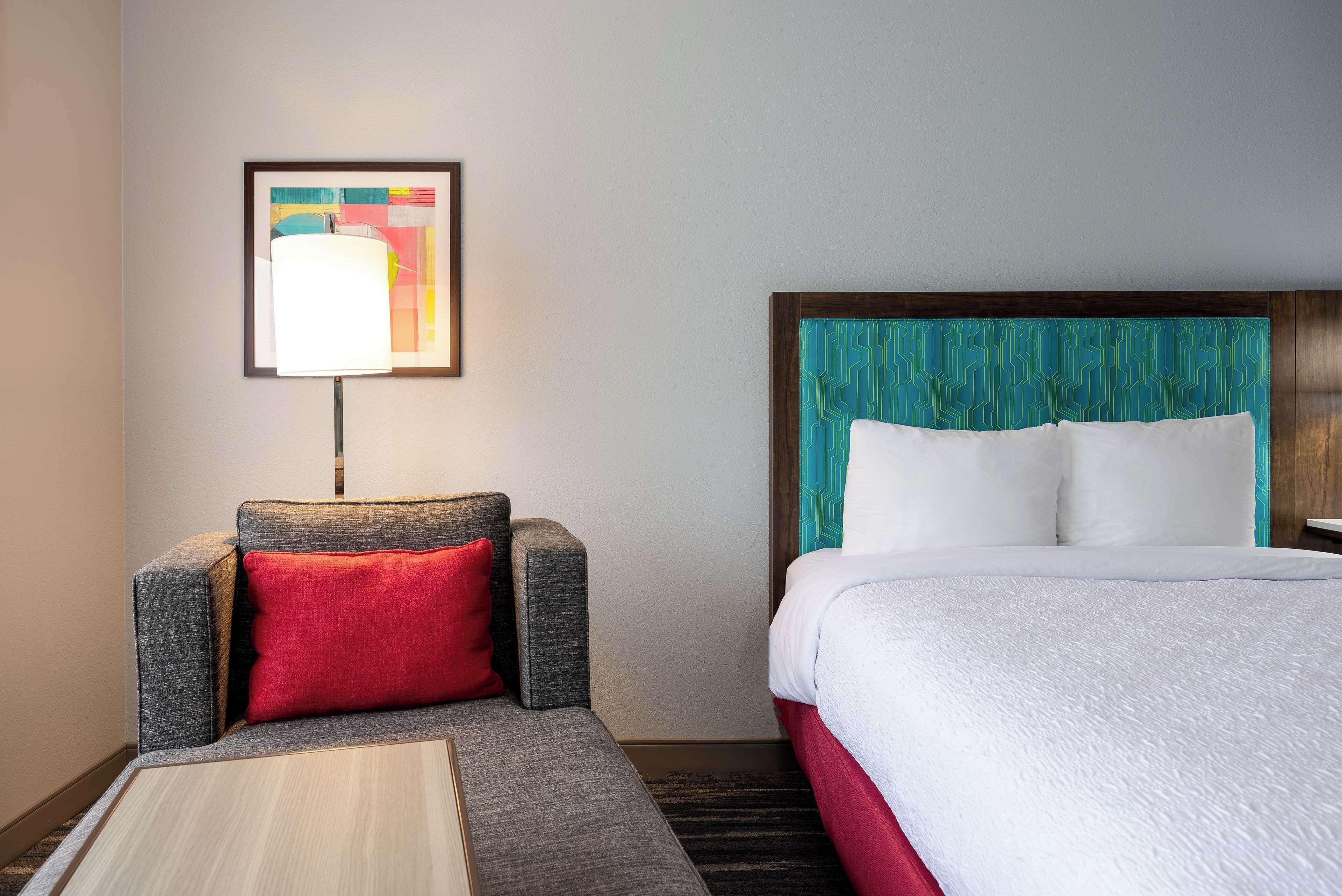 Quarto de hotel com cama, mesa de cabeceira, luminária e uma obra de arte abstrata emoldurada na parede.