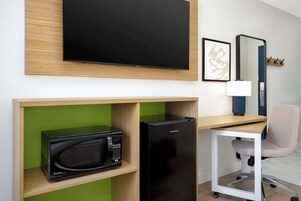 Quarto de hotel moderno com televisão de tela plana na parede, micro-ondas e frigorífico numa prateleira de madeira, uma secretária com cadeira e um espelho com lâmpada.