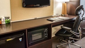 Quarto de hotel com televisão de tela plana na parede, uma mesa com luminária, micro-ondas, cadeira de escritório preta e um minibar com lanches.