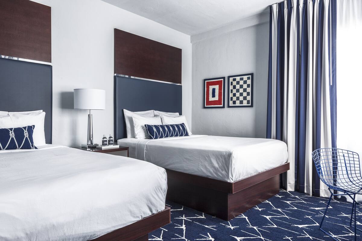 Quarto de hotel com duas camas, uma escrivaninha, uma cadeira e um detalhe decorativo na parede com padrão xadrez.