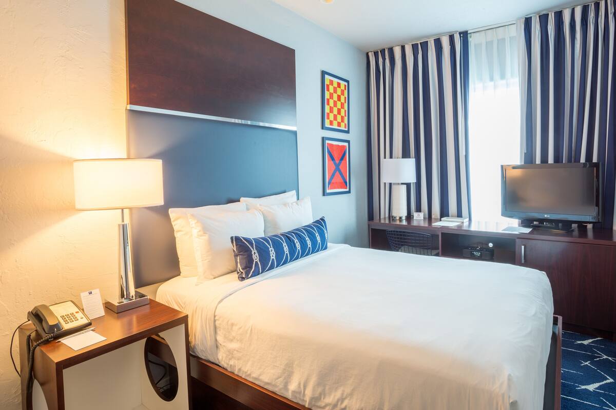 Quarto de hotel com cama, luminária de cabeceira, escrivaninha com telefone e televisão.