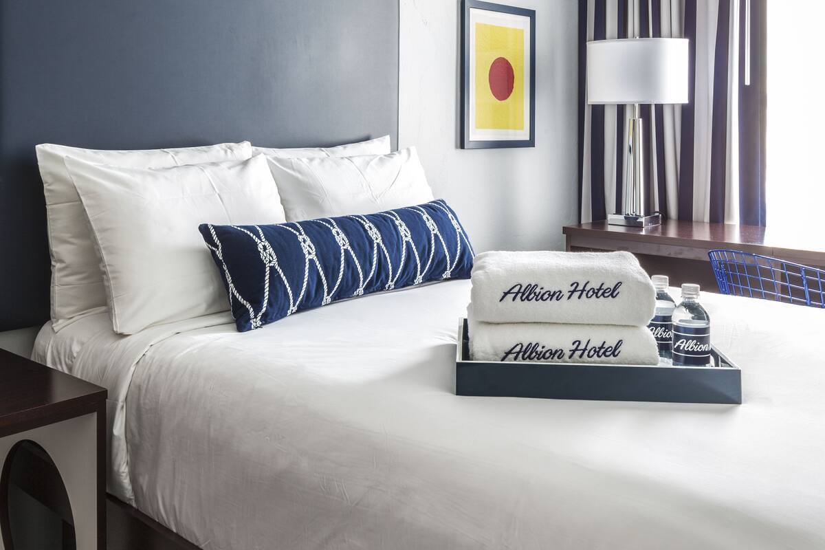 Quarto de hotel com cama arrumada, mesa de cabeceira com abajur e bandeja com toalhas e garrafas de água.