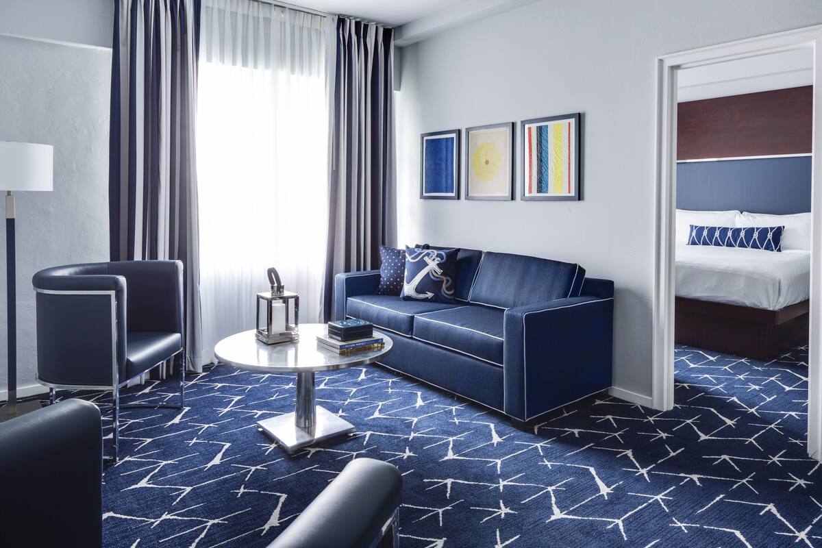 Quarto de hotel moderno com um sofá azul, uma mesa de centro redonda, uma cama com cabeceira e um tapete estampado.