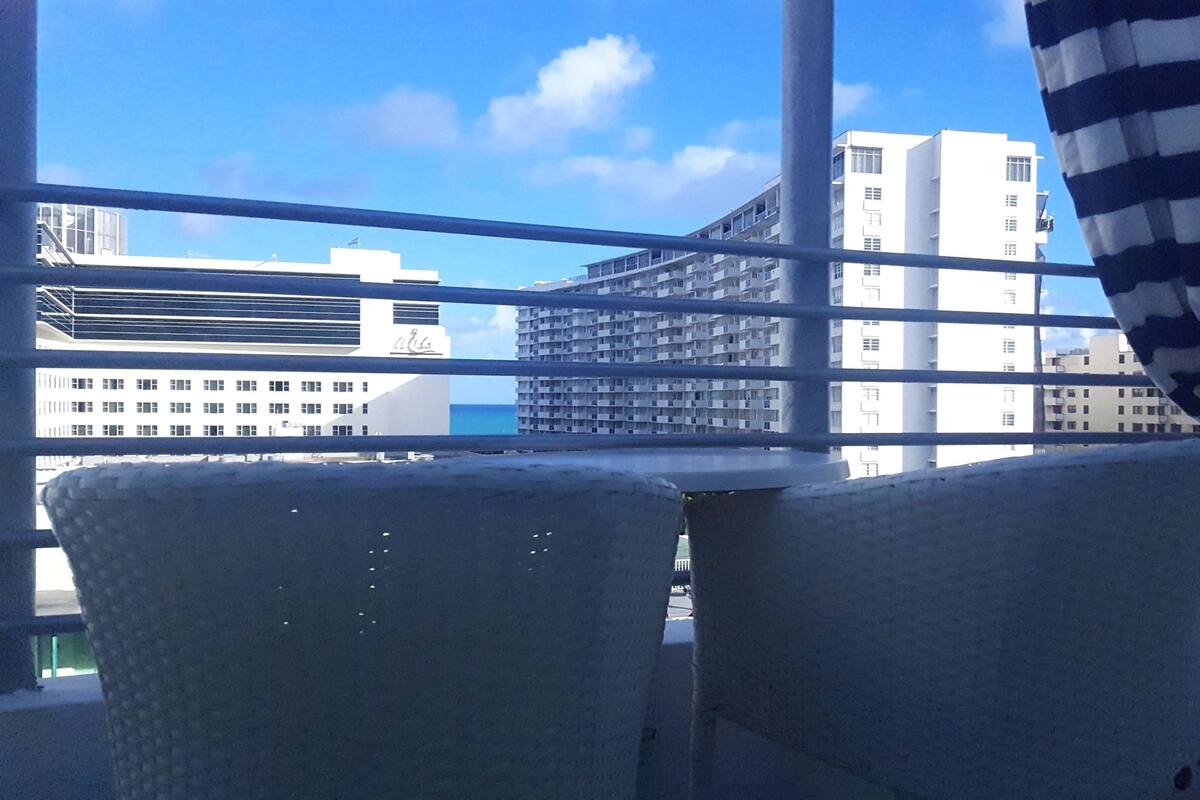 Uma varanda com duas cadeiras de vime, uma cortina listrada e vista para edifícios altos e o oceano.