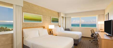 Quarto de hotel com duas camas, uma escrivaninha, uma televisão e vista para o oceano.