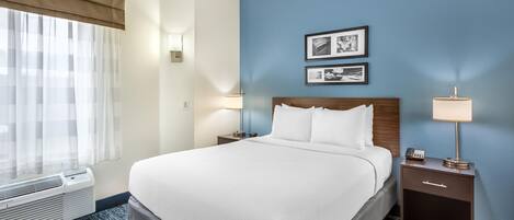Quarto de hotel com uma cama grande, duas mesas de cabeceira com luminárias, um ar condicionado fixado na parede e uma janela com cortinas leves.