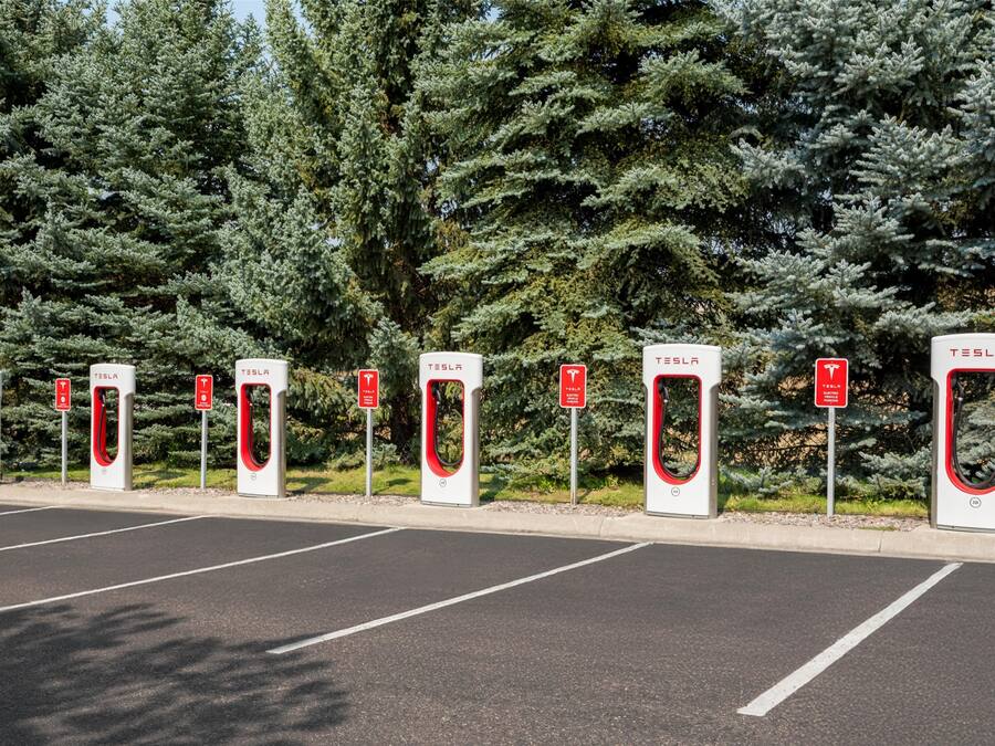 Fileira de estações de carregamento para veículos elétricos Tesla, com sinalização vermelha, ao fundo árvores perenes.