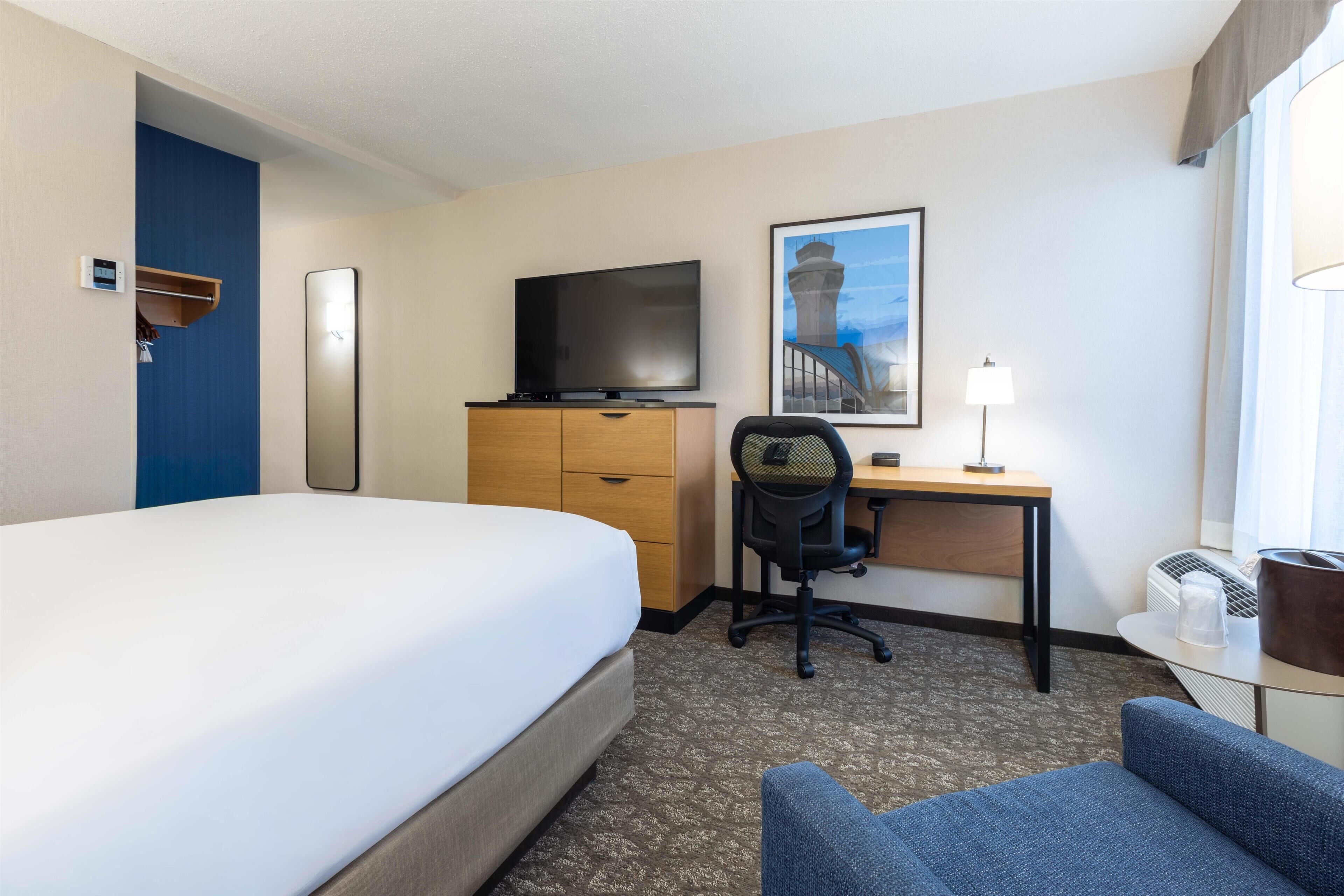 Quarto de hotel com cama, escrivaninha, cadeira, televisão e um quadro na parede.