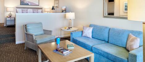 Quarto de hotel com um sofá azul, uma cadeira de vime, uma mesa de centro de madeira e uma mesinha de cabeceira com uma luminária.