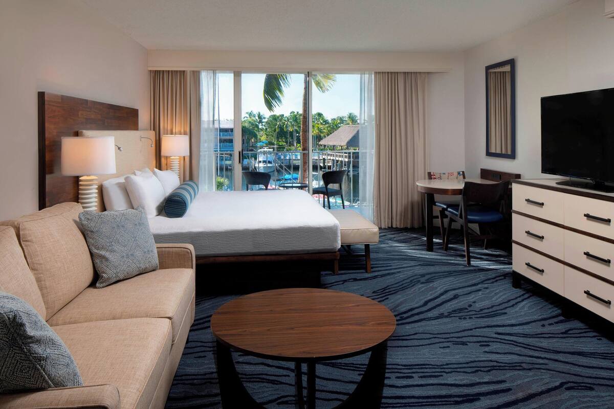 Quarto de hotel com cama, sofá, televisão, escrivaninha e vista para a piscina e palmeiras.