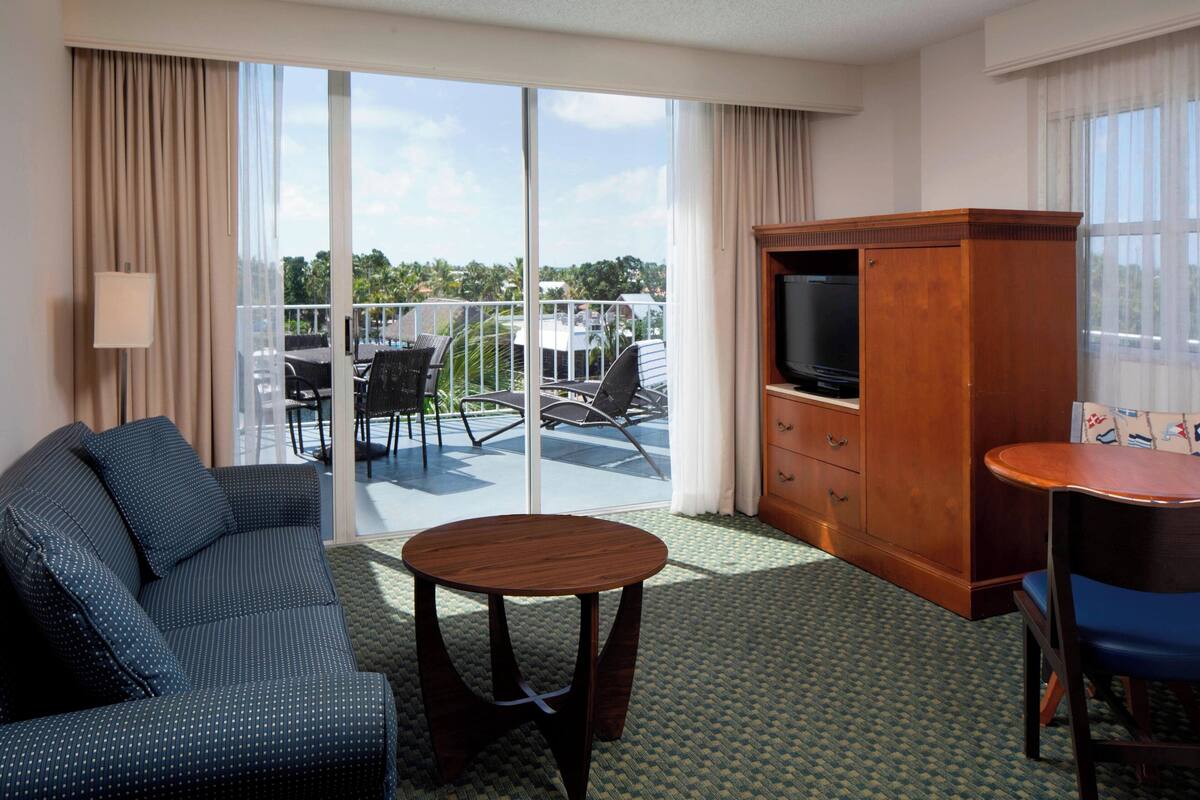 Quarto de hotel com sofá, uma mesa redonda pequena, um aparador de TV e varanda com mobiliário exterior.