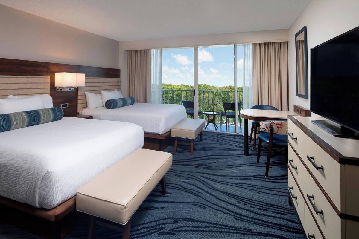 Quarto de hotel com duas camas, uma escrivaninha, televisão e varanda com vista.