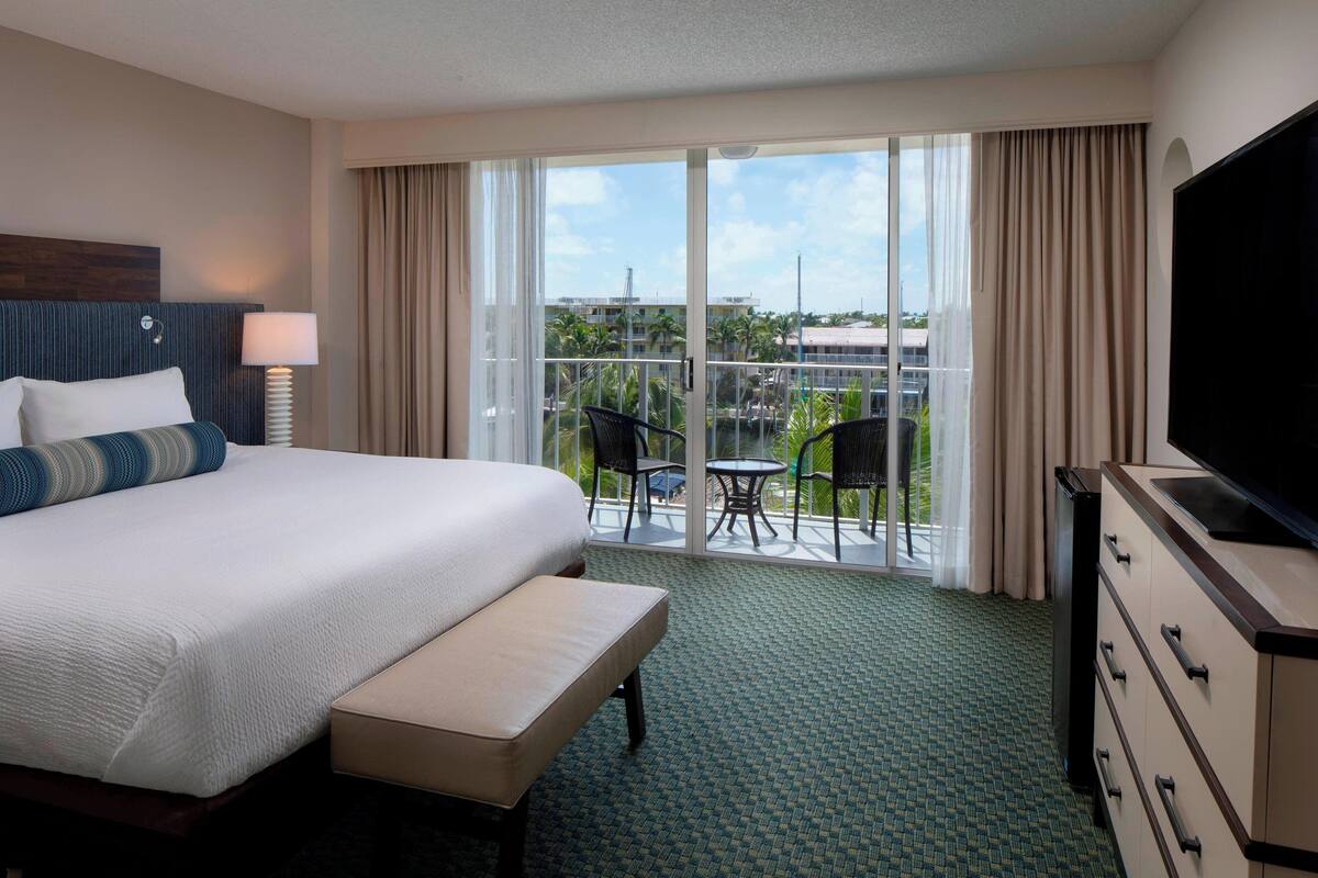 Quarto de hotel com uma cama grande, televisão, escrivaninha e varanda com cadeiras e mesa.