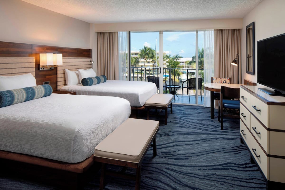 Quarto de hotel com duas camas, uma mesa de trabalho, televisão e varanda com vista para palmeiras e edifícios.